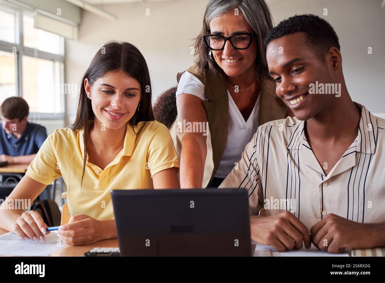 Smiling female teacher helping two multiracial college students using laptop to study in ...