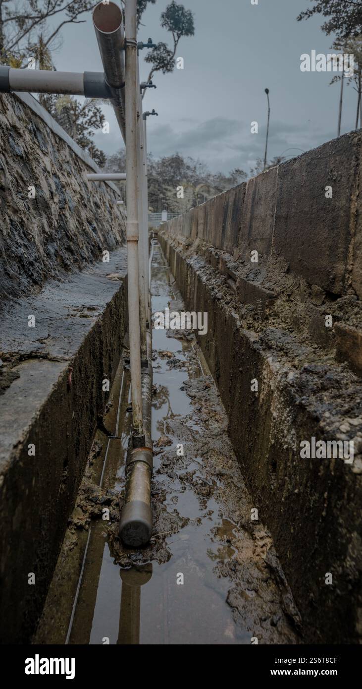 A narrow, muddy drainage canal with exposed pipes running along its ...