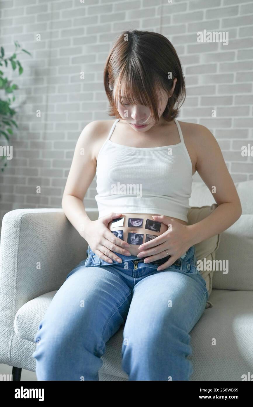 A Japanese pregnant woman in her 20s, dressed to show her stomach, sits on a sofa in her ...