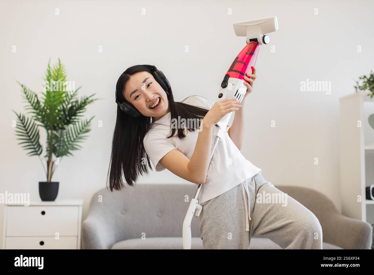 Asian young woman enjoying cleaning with headphones and vacuum. Happy and playful atmosphere in ...