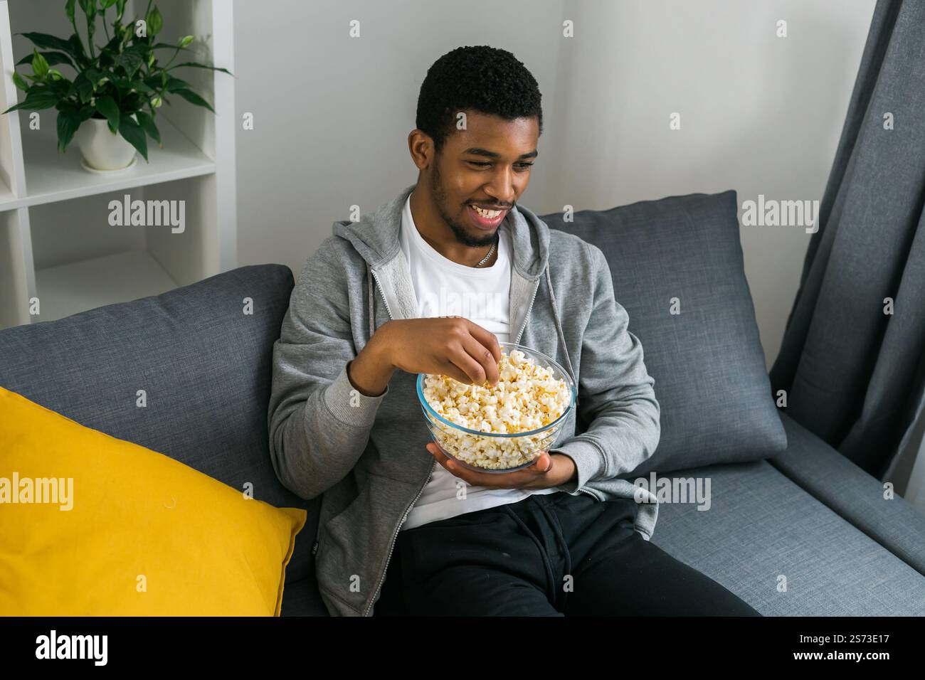African American man sits alone on the couch and enjoying popcorn while watching television and ...