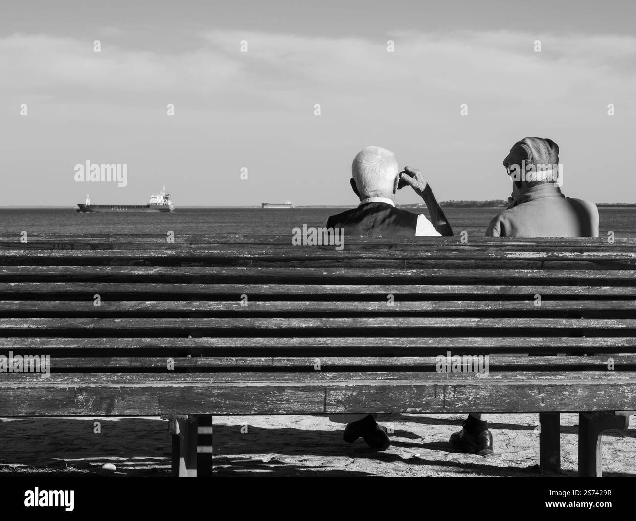 Lisbon, Portugal. 2023. Two men sit on a waterside bench as boats and ...