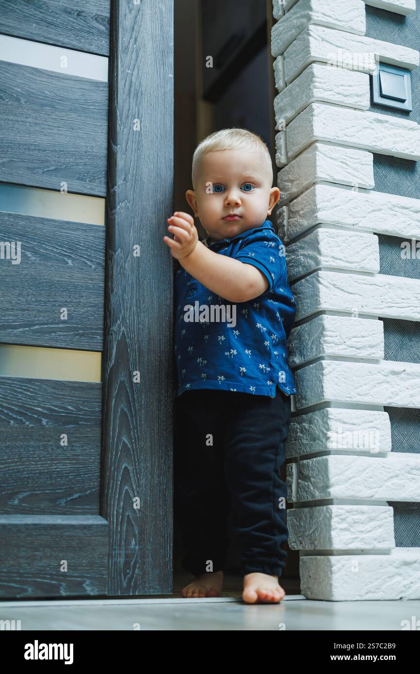 Blond toddler boy at home peeks into the room through a crack in the ...