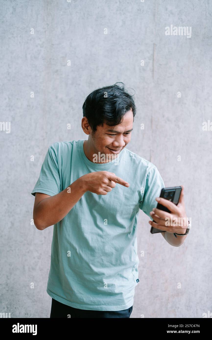 Asian man excitedly pointing at his smartphone against a gray ...