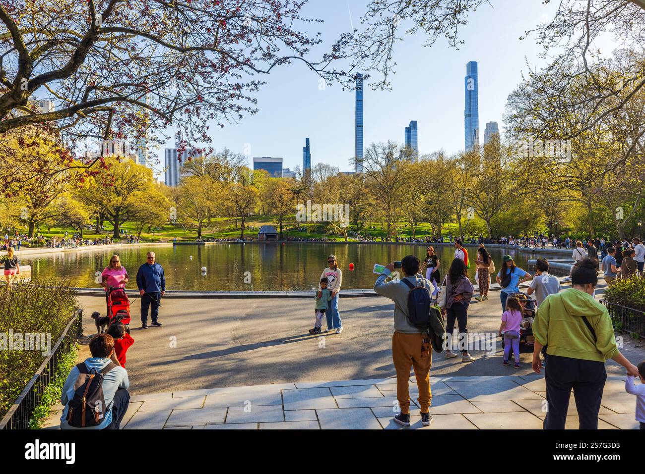 People enjoying warm spring day at Central Park near pond with city ...