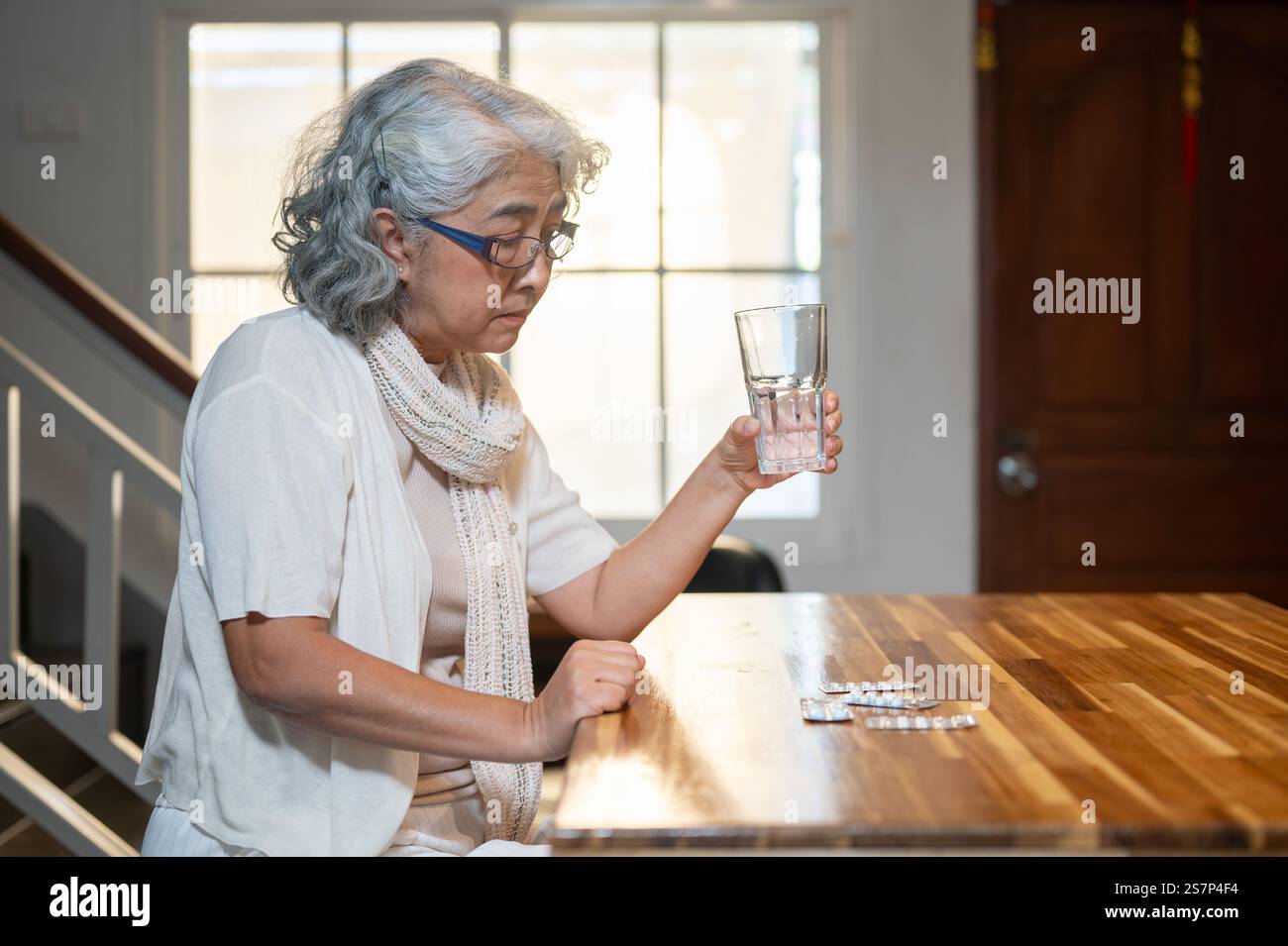 An unwell, elderly Asian woman drinks water while taking medicine at a ...