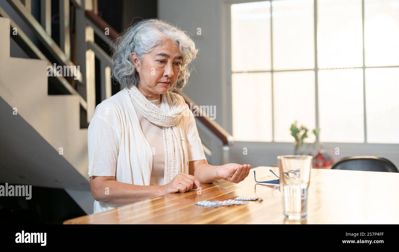 An unwell, elderly Asian woman takes medicine at a dining table ...