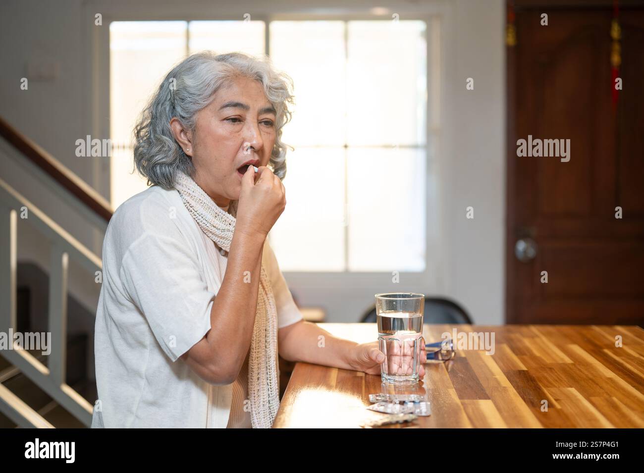 An unwell, elderly Asian woman takes medicine at a dining table ...