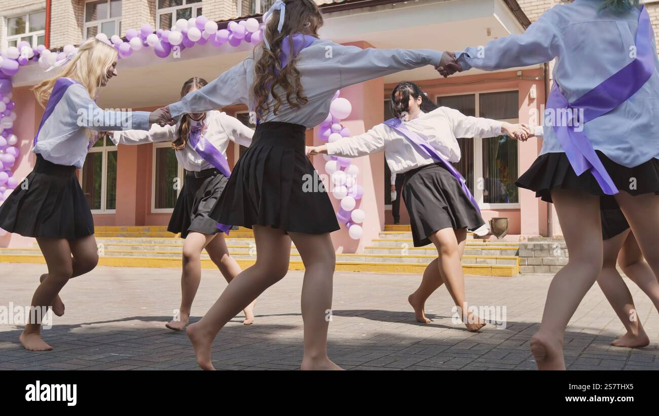 Female graduates dancing barefoot in schoolyard, celebrating their last ...