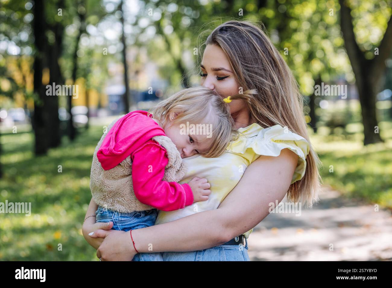 Nurturing mother comforting crying child, embracing closely near leafy trees on bright summer ...