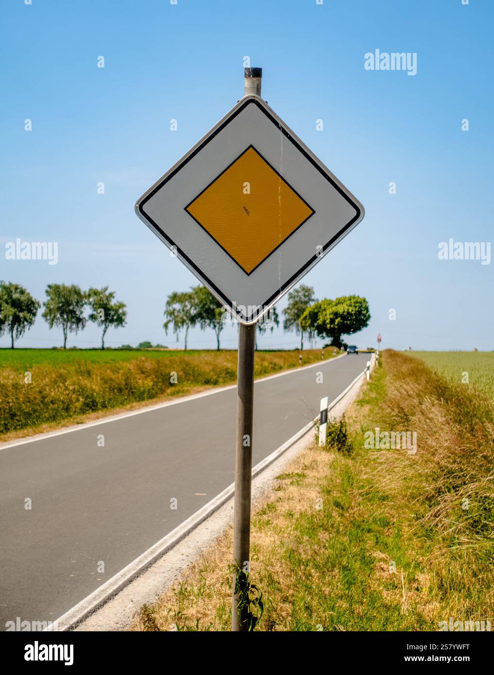 A yellow and white sign is on a road with a green field in the ...