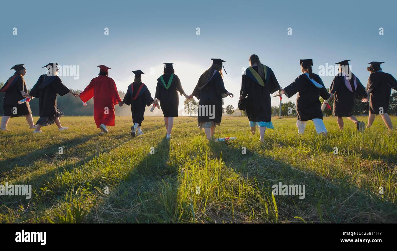 Group of graduates in robes walking hand in hand into the sunset in a ...