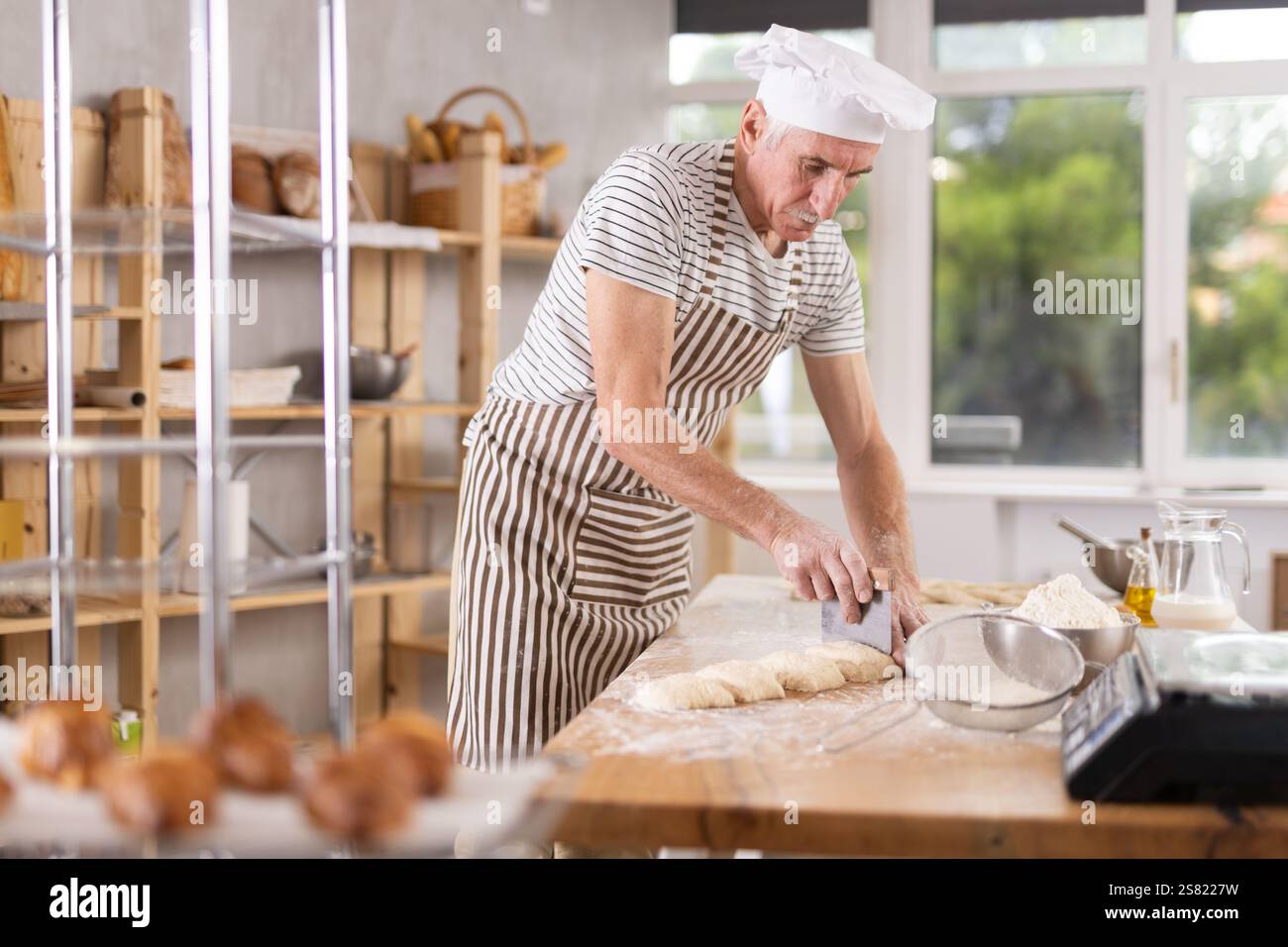 Elderly experienced male baker using knife to cut raw dough to make ...