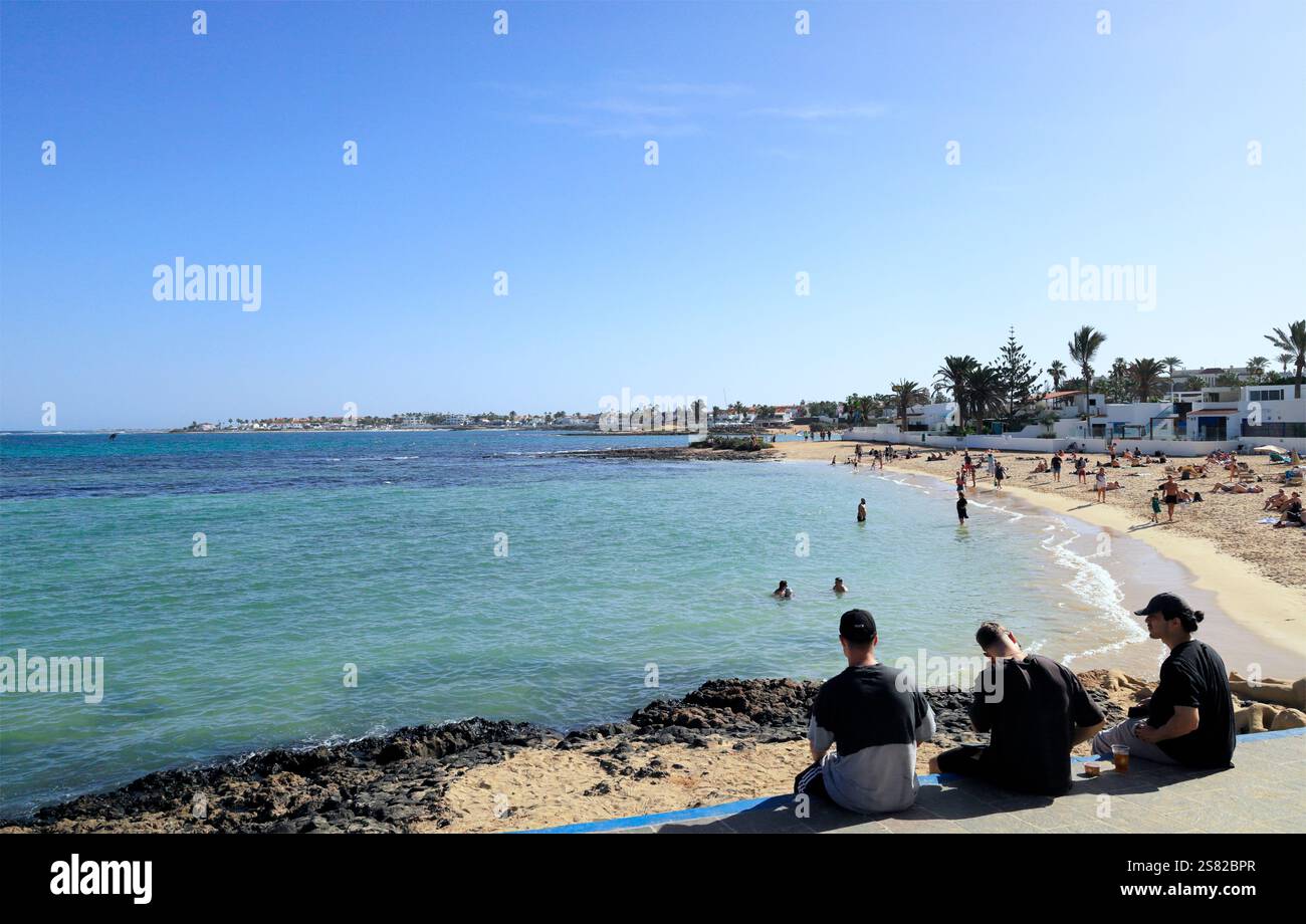 Three young men sit relaxing by the beach at Corralejo, Fuerteventura ...