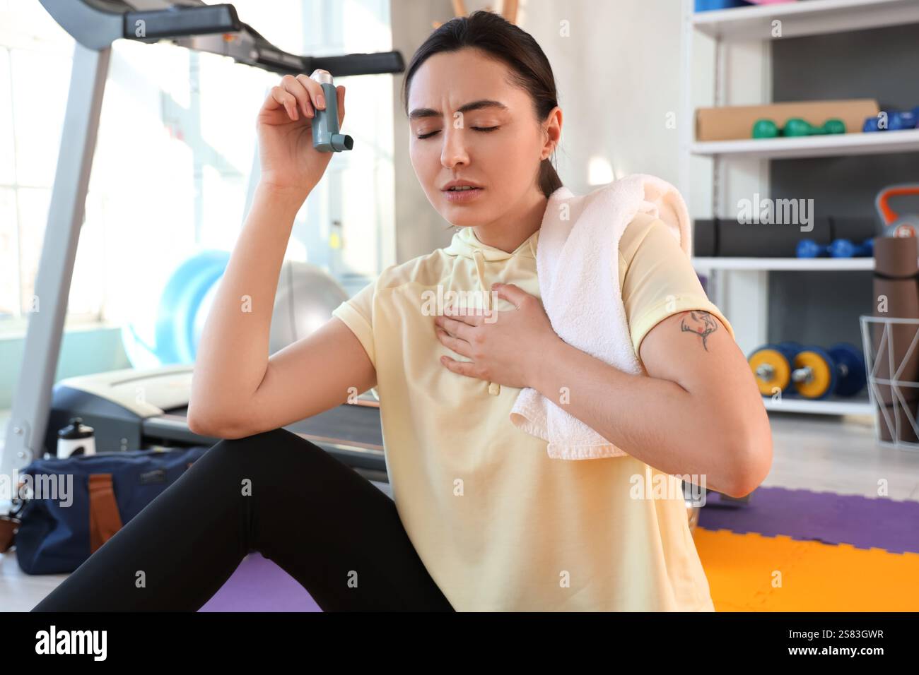 Sporty young woman with inhaler and towel having asthma attack in gym ...