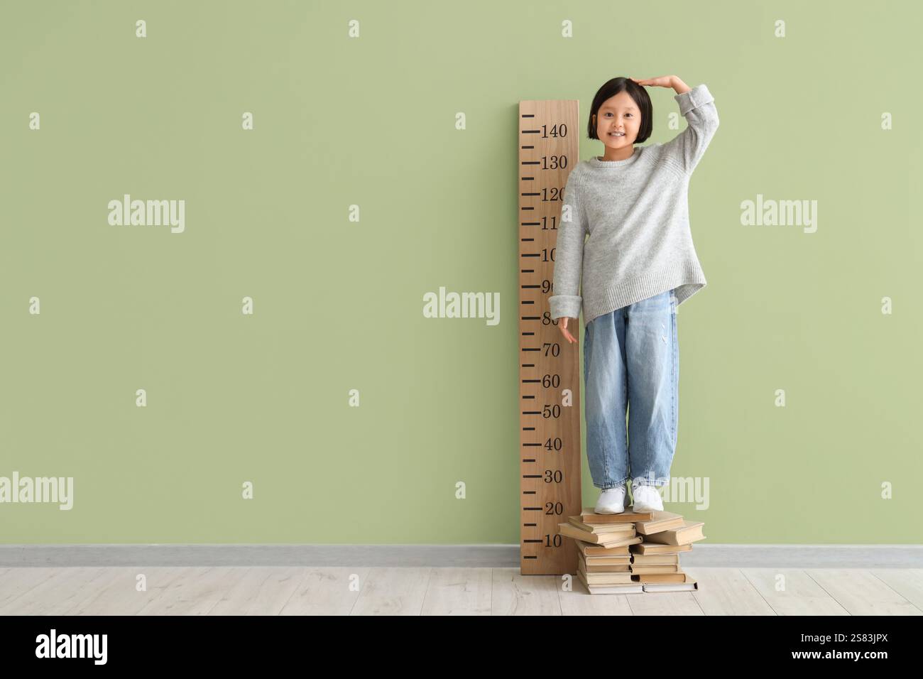 Cute Asian girl on books measuring height with stadiometer near green ...