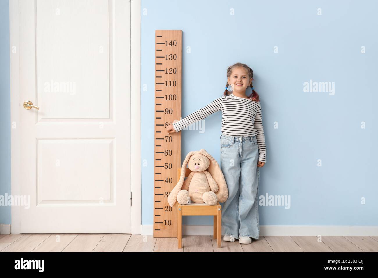 Little girl measuring toy's height on chair near stadiometer at home ...
