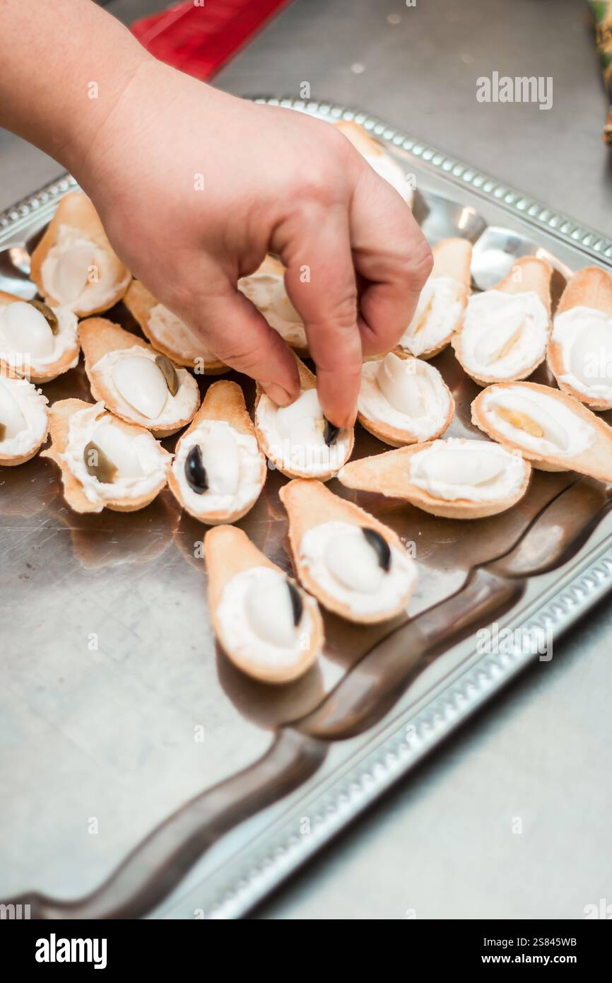 A hand places a black olive on a pastry shell filled with creamy white ...