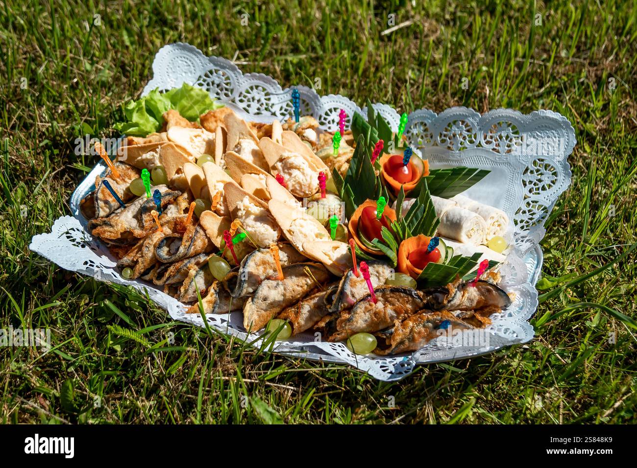 An ornate platter on grass features grilled fish, bread rolls ...