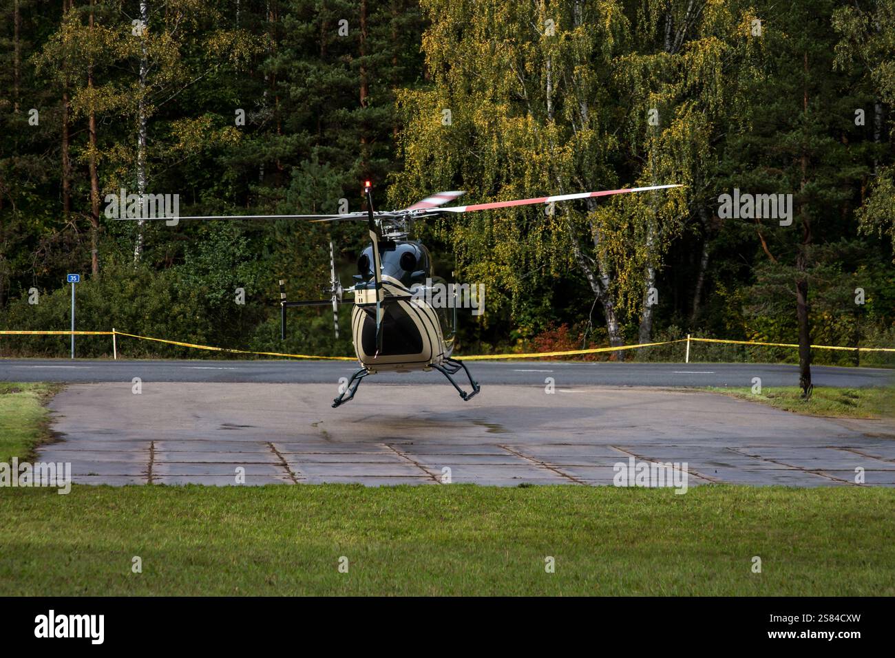 A helicopter lands on a paved helipad bordered by grass and dense ...