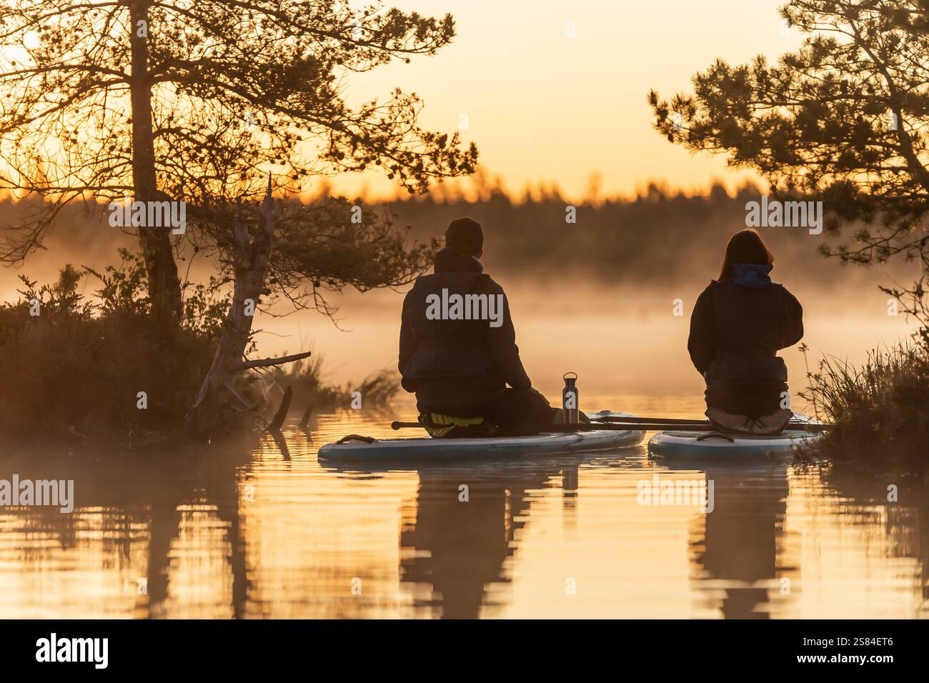 Two paddleboarders sit on a calm waterway with mist rising, golden ...