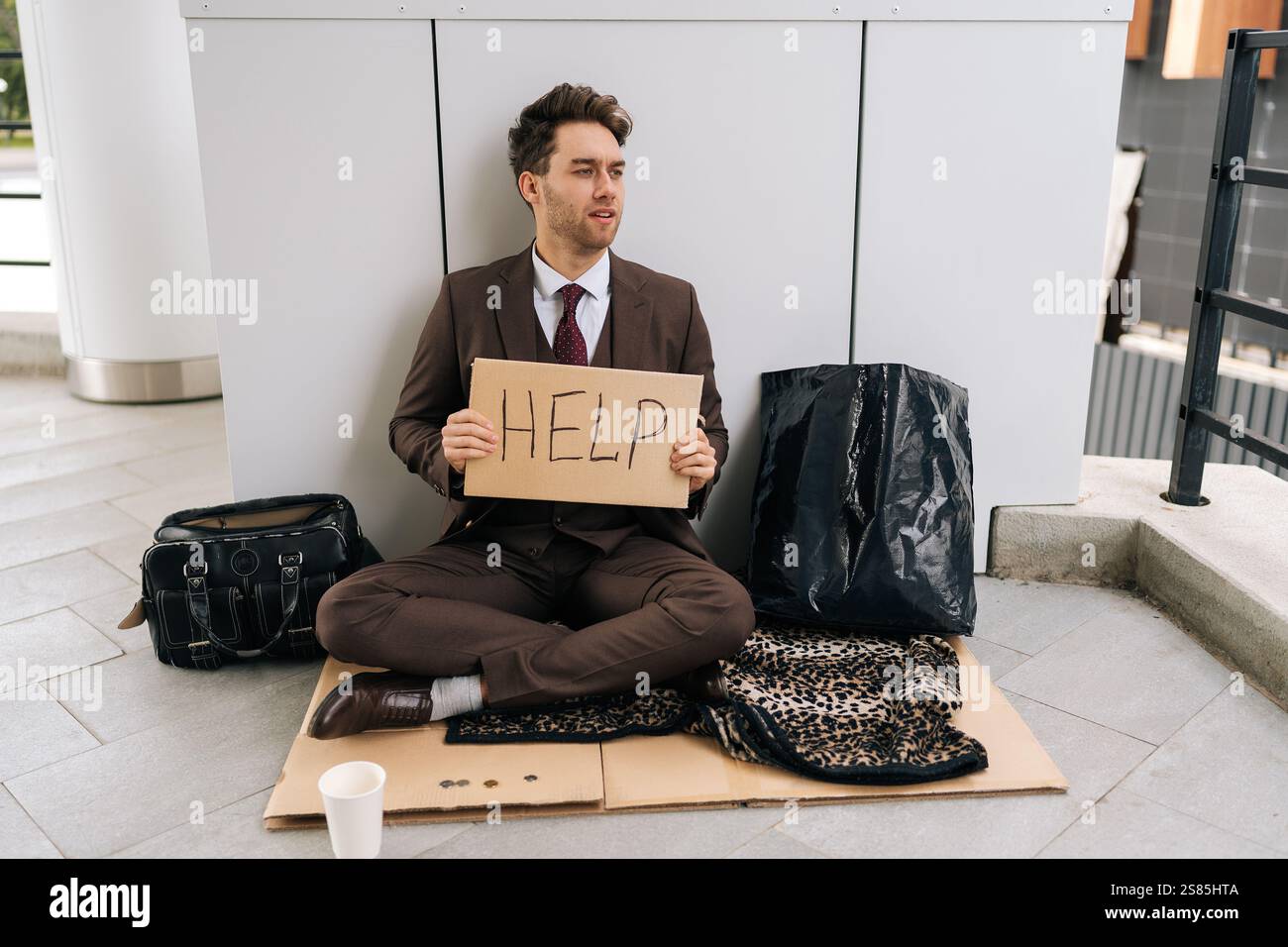 Homeless millennial businessman in suit sitting on city street holding ...