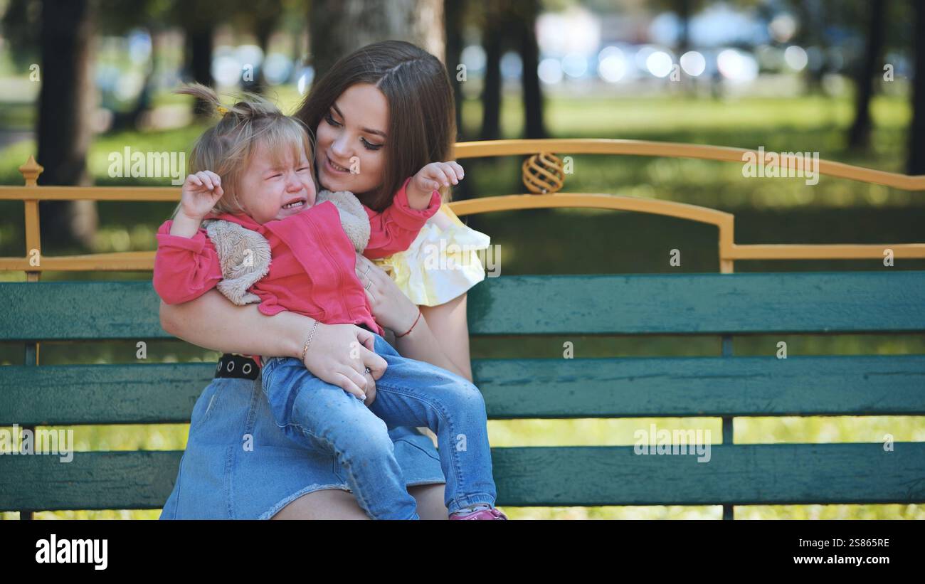 Caring mother soothing distressed child, seated on park bench under ...