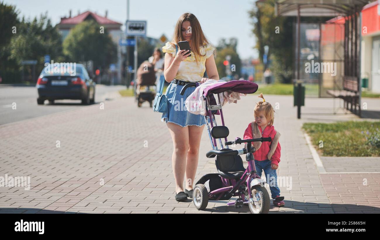 Urban mom checking smartphone, pushing tricycle riding daughter along ...