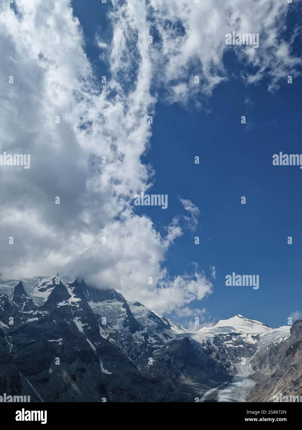 Majestic snow-covered mountain peak under a blue sky, with clouds forming at the summit ...