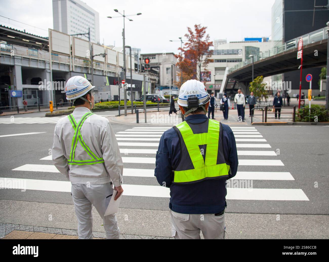 Two workmen with hard hats and safety gear waiting to cross a road in ...