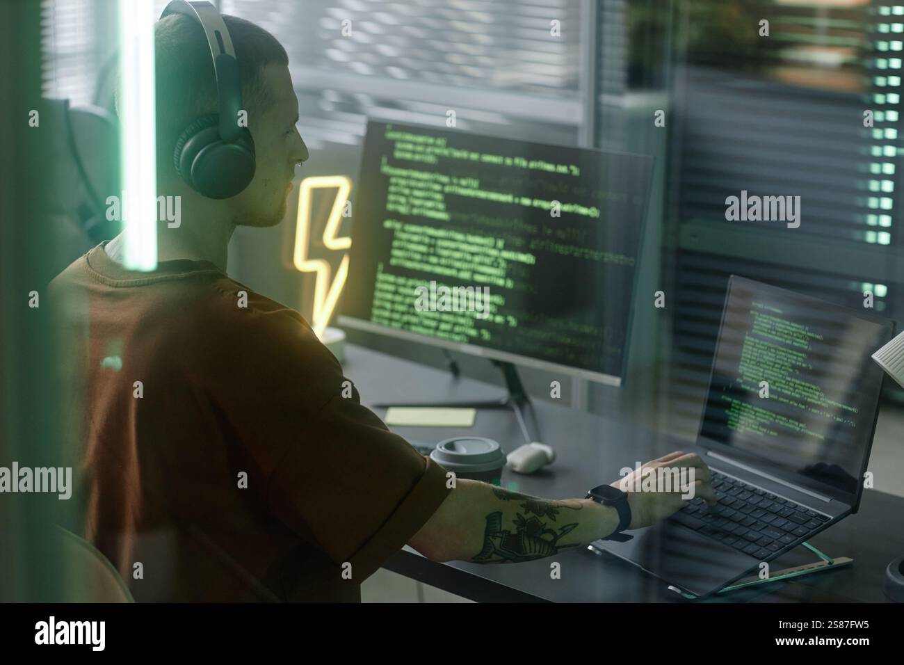 Shot through glass wall of male senior developer in headphones coding ...