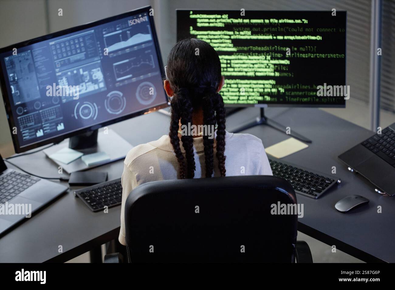 Rear view of female programmer with braids hairstyle coding at desk with several computer ...