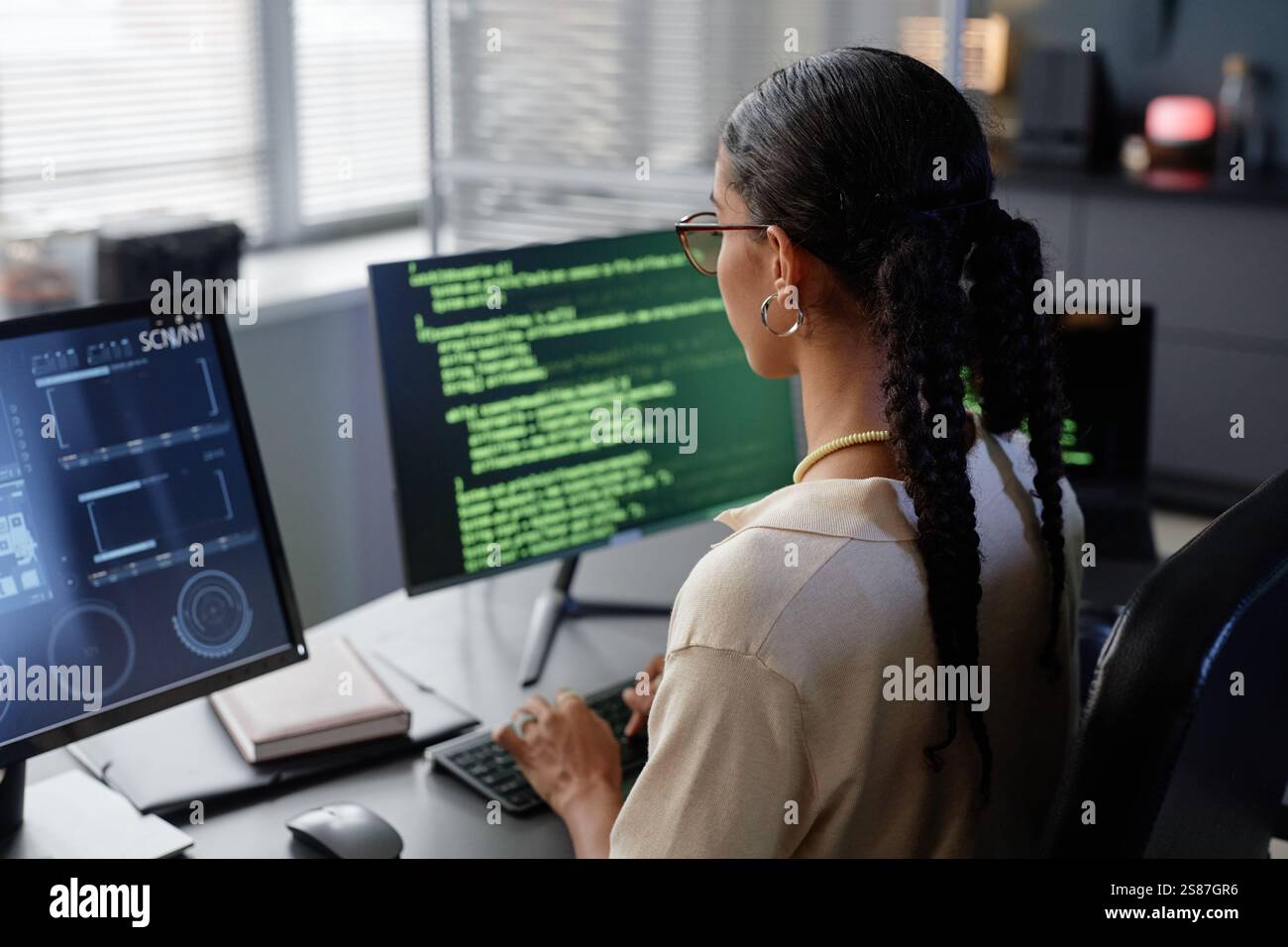 Rear view of Middle Eastern female programmer with glasses reading multiple computer screens ...