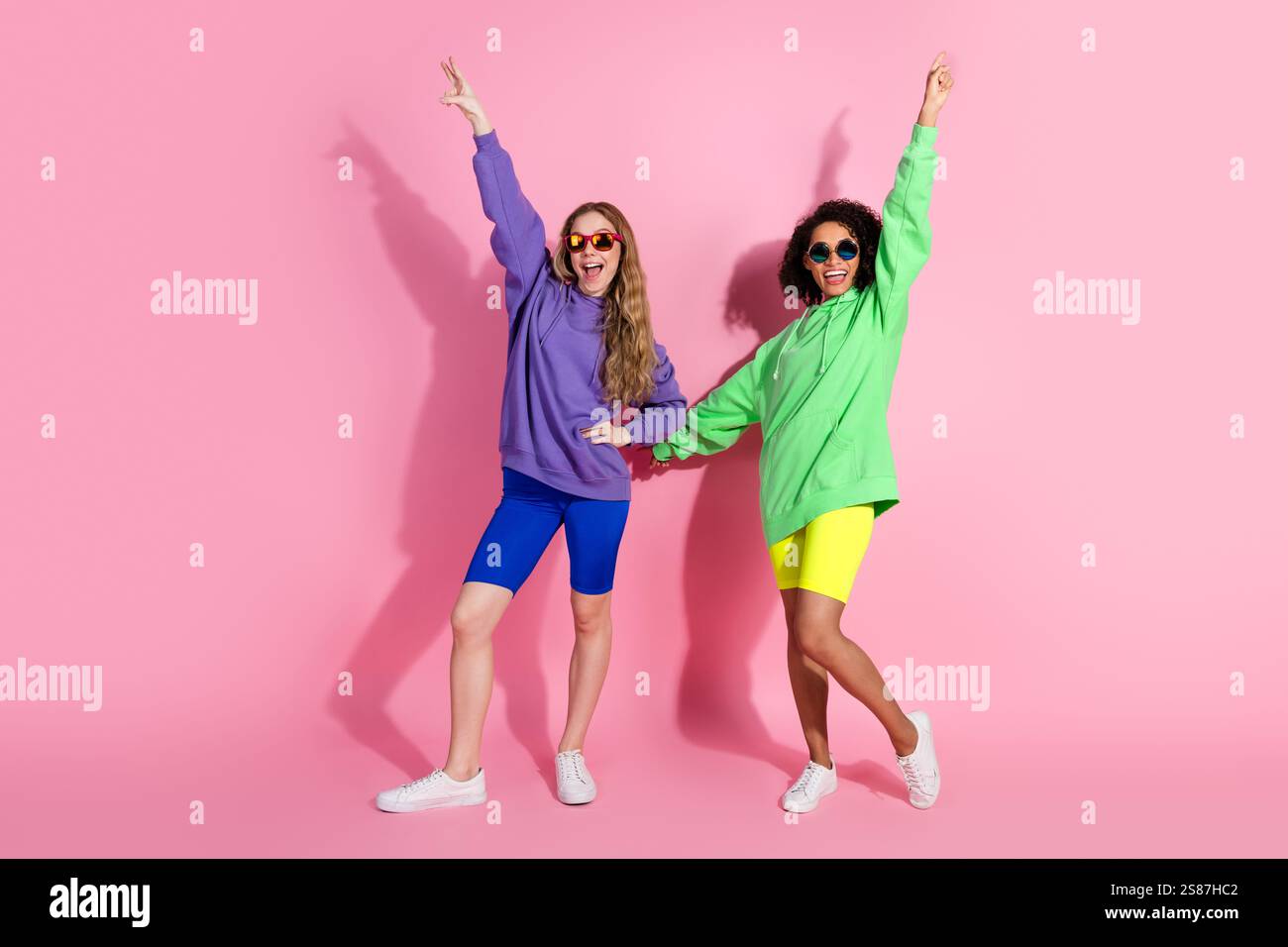 Two joyful young women in colorful outfits dancing on pink background ...