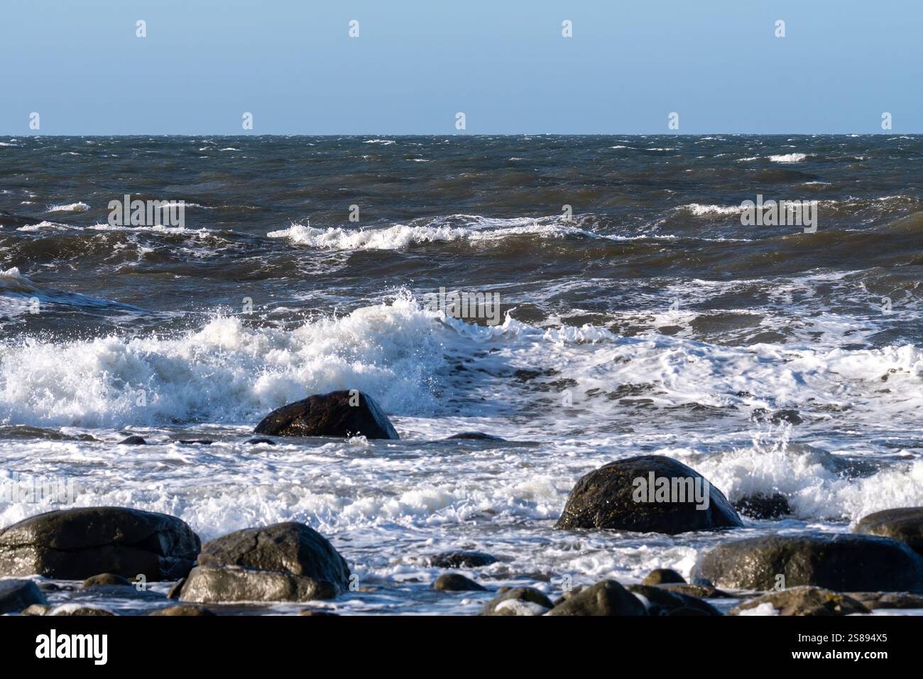 Choppy waves collide with large, dark rocks along the coastal shore ...
