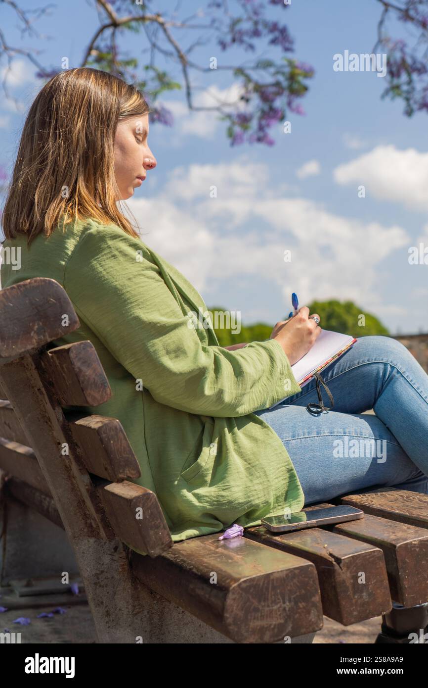 A vertical shot of a young woman sitting on a bench in a park, writing ...