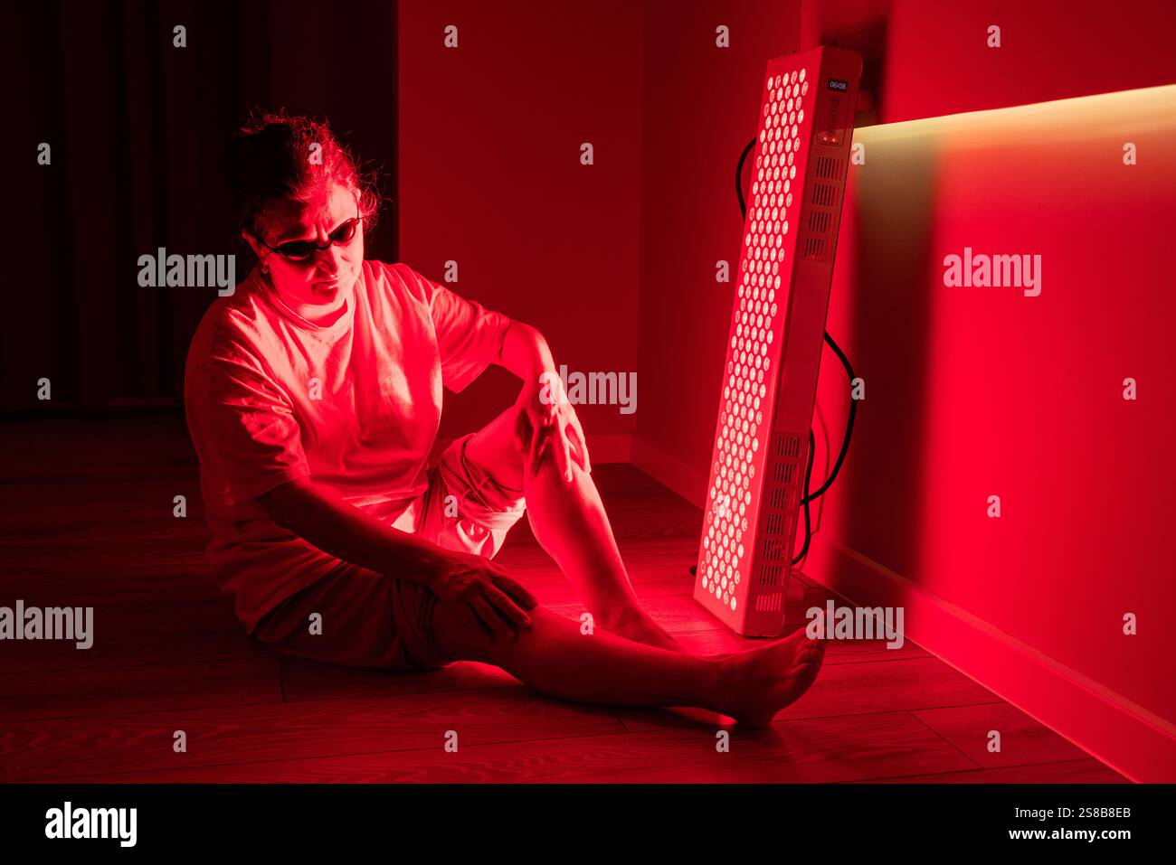 Elderly woman seated using a red light therapy panel on her sore leg ...