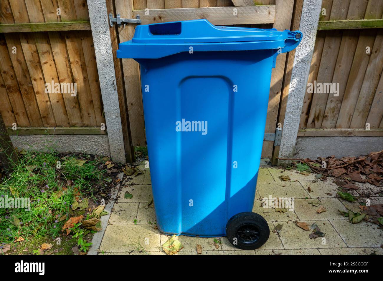 A blue wheelie bin is positioned against a wooden fence in a backyard ...