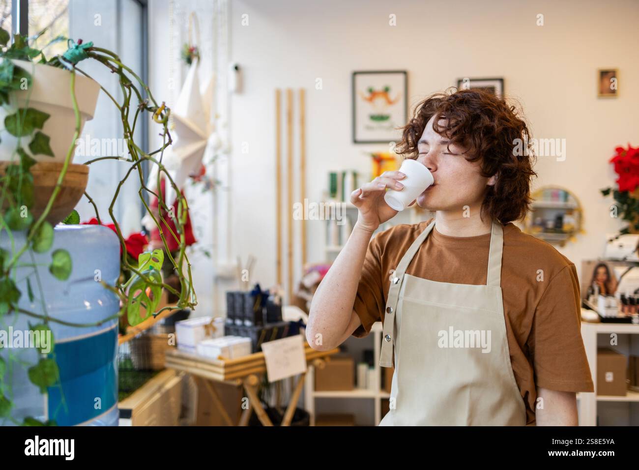 A young shop assistant in an apron takes a break, drinking water from a ...