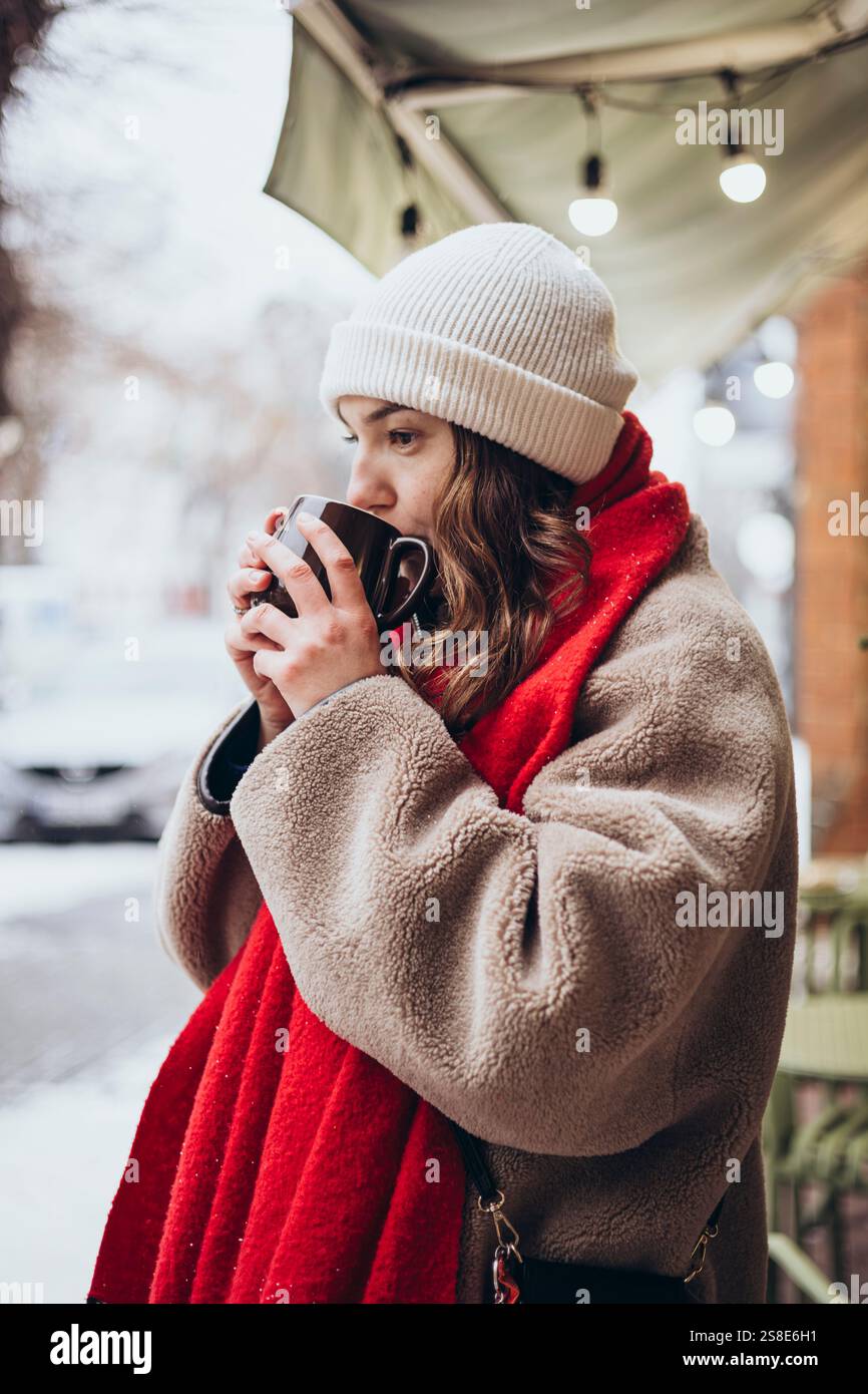 A young thoughtful woman sips a warm beverage outdoors in winter ...