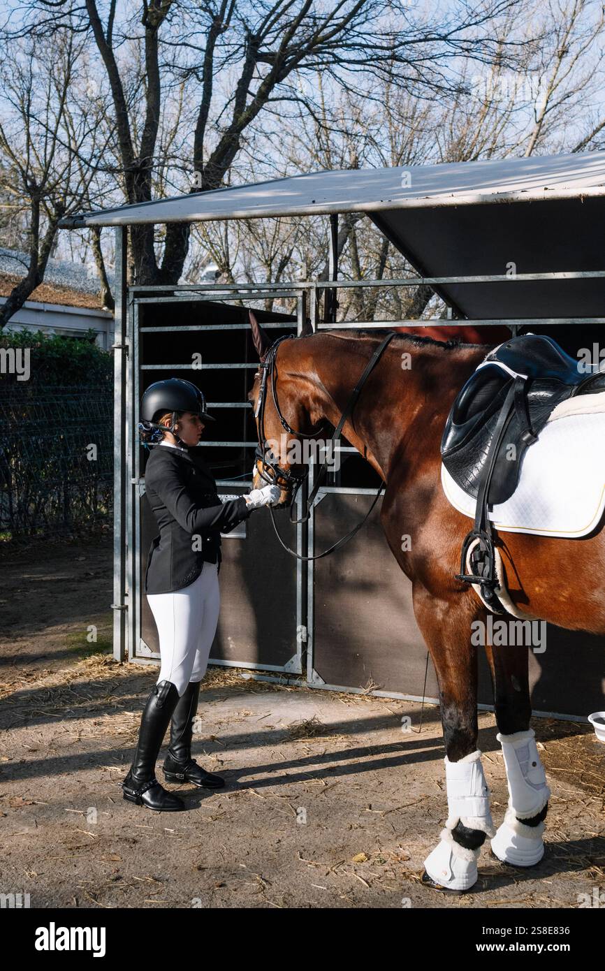 A teenage girl in a classical dressage attire is adjusting the bridle ...