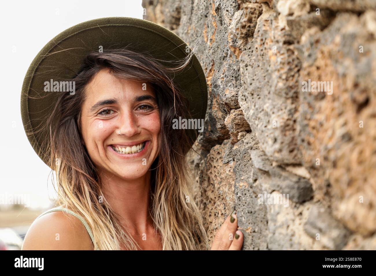 A woman wearing a hat smiles brightly while standing against a textured ...