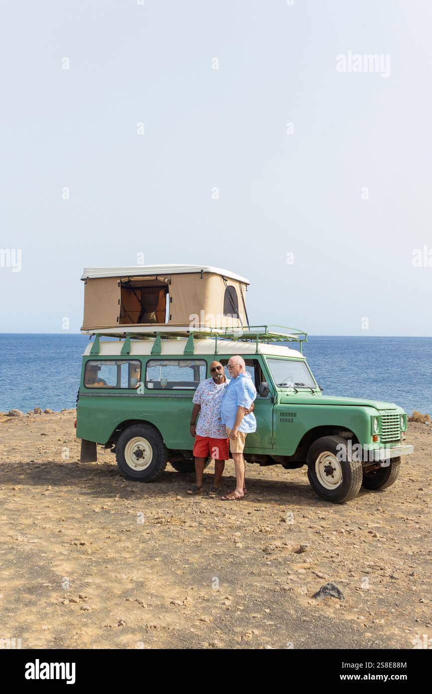 A joyful gay senior couple embraces beside an off-road vehicle during a ...