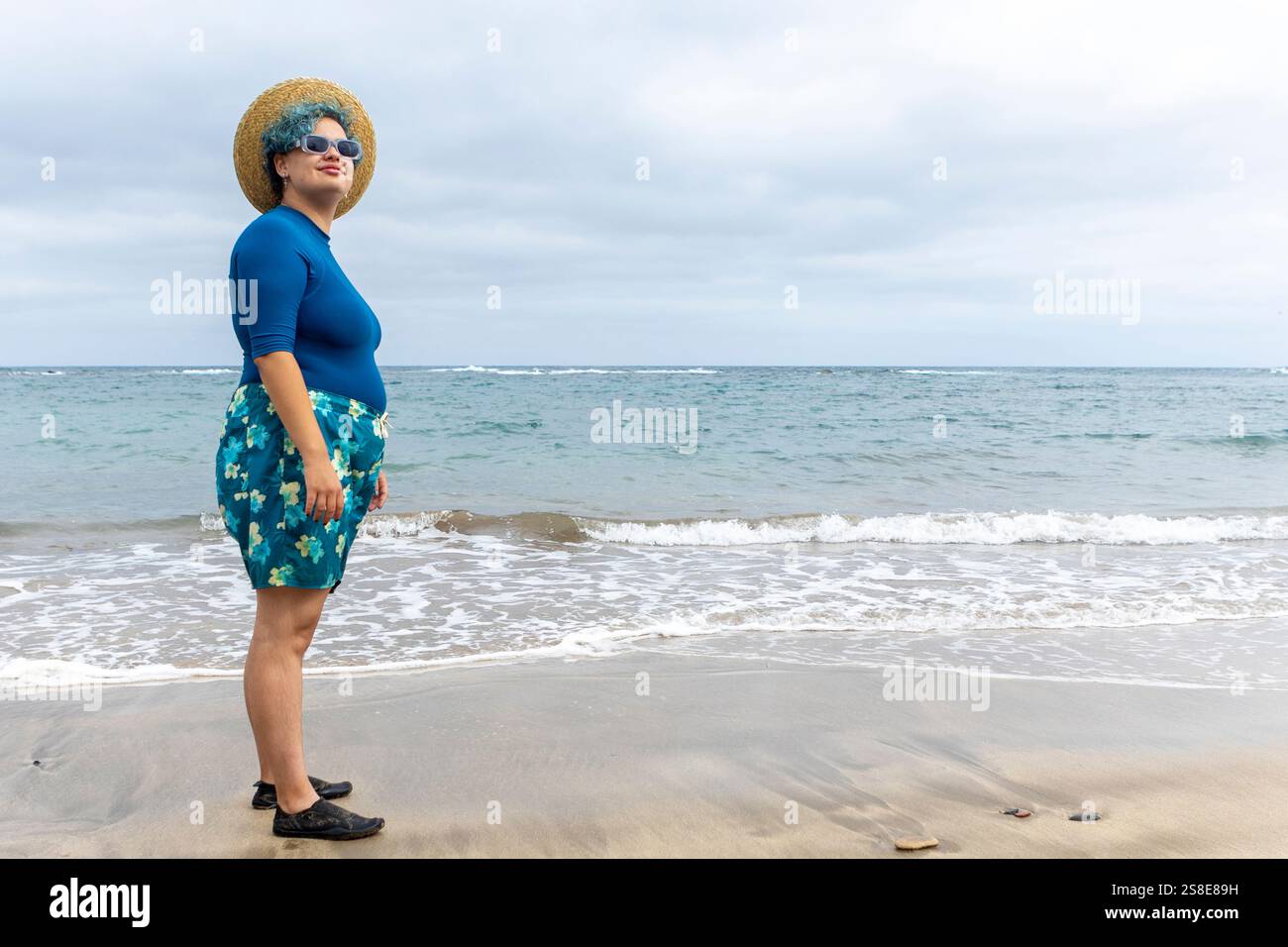 A transgender man, stands on a beach looking towards the sea. Wearing a ...