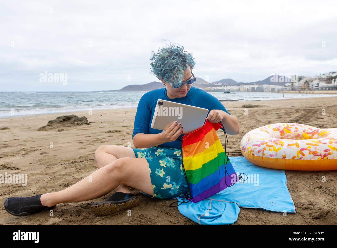 A transgender man with blue hair sitting on a beach, putting a tablet ...