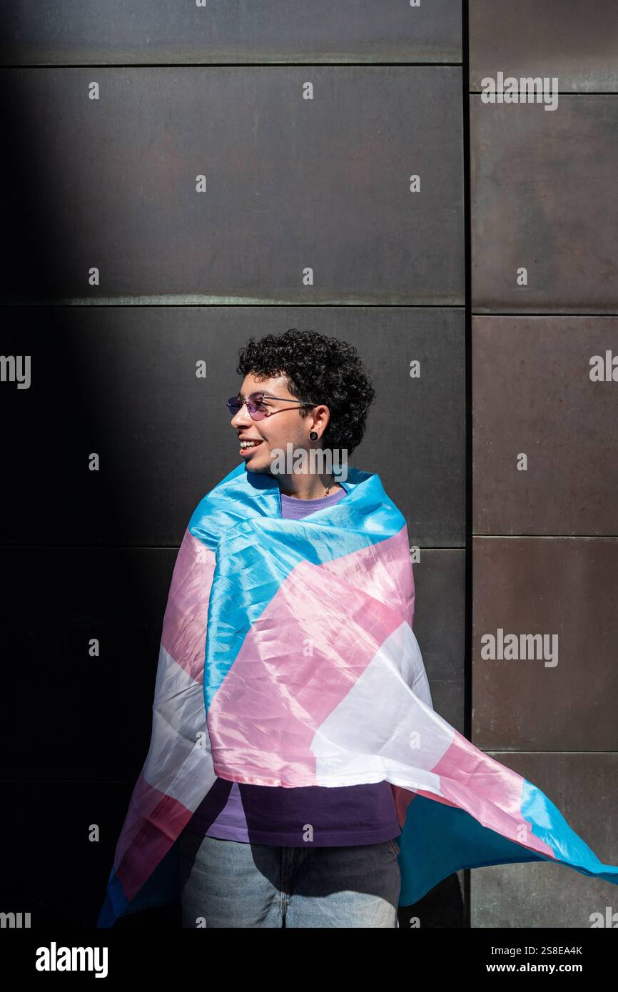 A LGBTQ+ person wrapped in a transgender pride flag smiles warmly ...