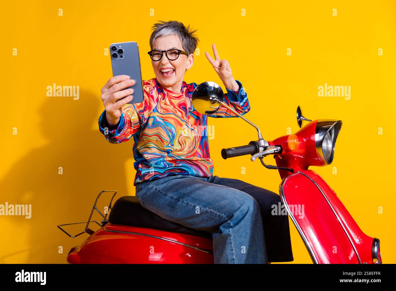 Portrait of pretty aged lady drive bike smart phone selfie v-sign isolated on yellow color ...