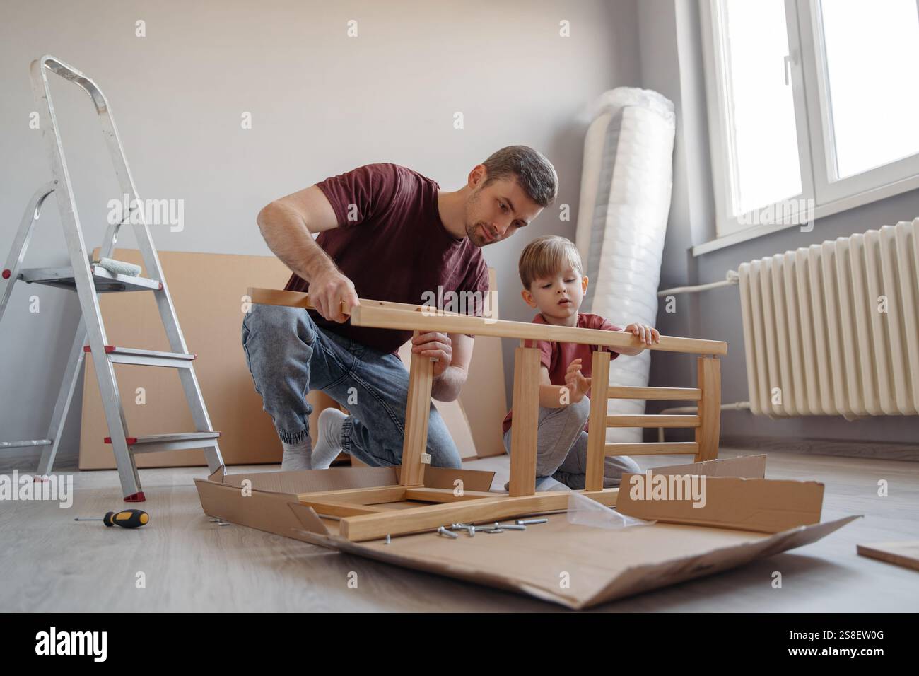 A father and his young son assembling wooden furniture at home, promoting teamwork, skill ...