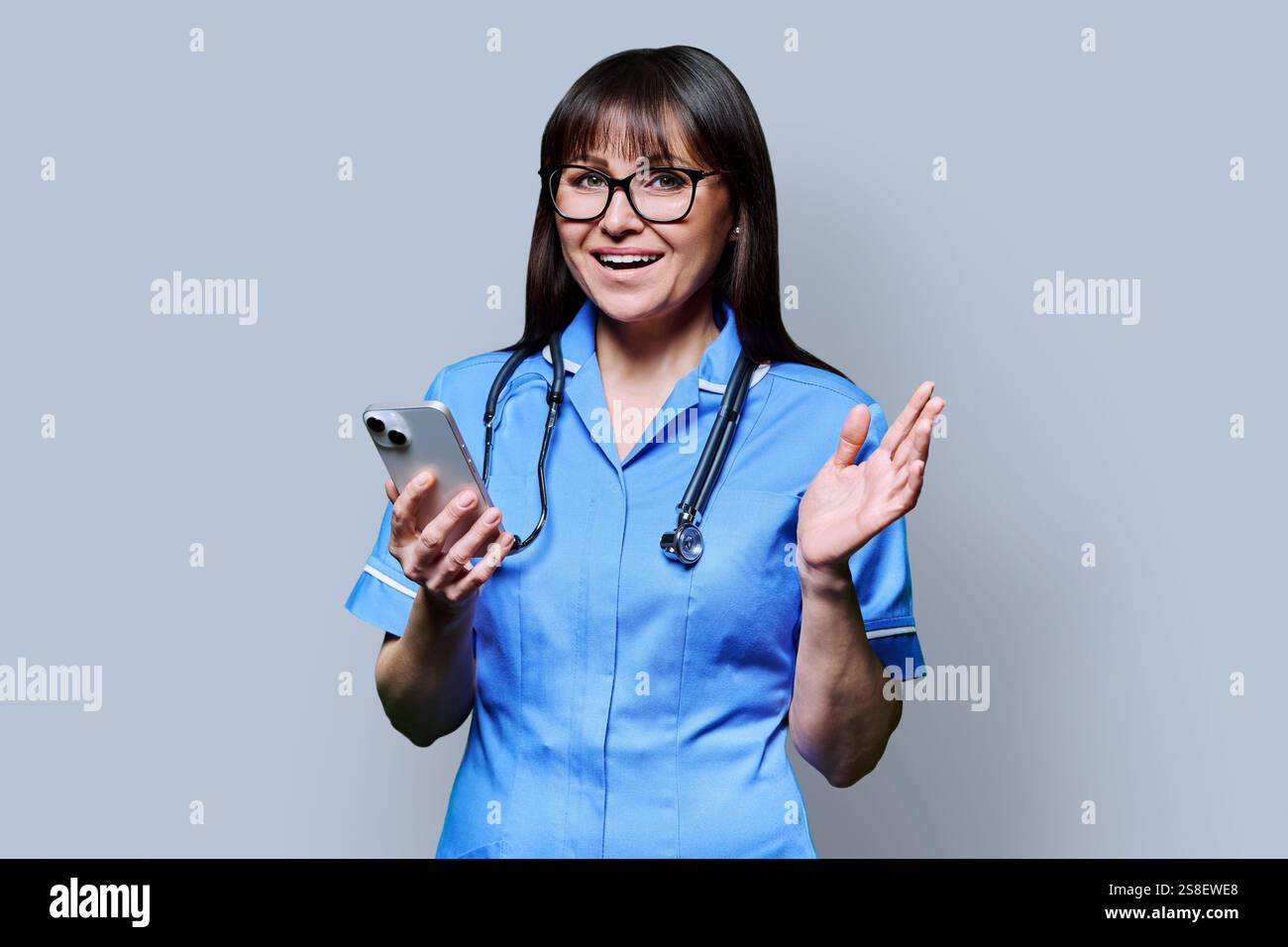 Portrait of smiling female nurse with smartphone, on grey studio ...