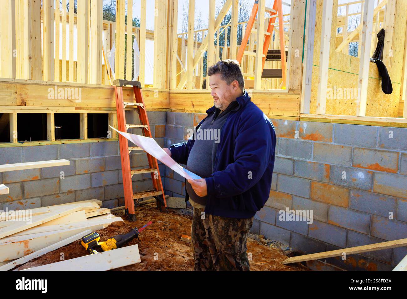 Construction worker stands on site studying blueprints at framing stage ...